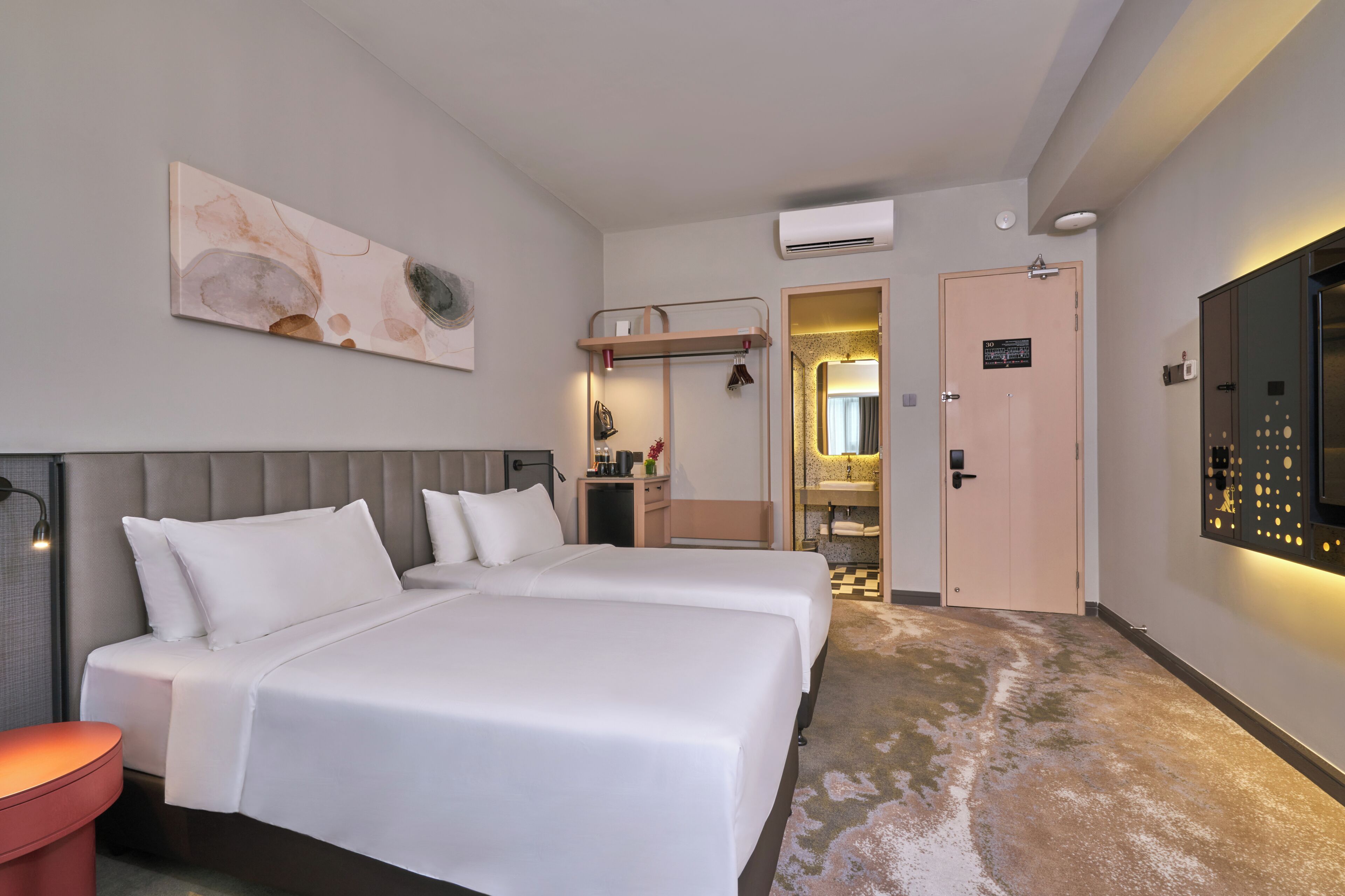 Quarto de hotel com duas camas, uma mesinha de cabeceira, TV na parede, ar condicionado e banheiro visível por uma porta aberta.