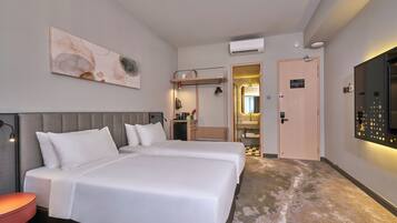 Quarto de hotel com duas camas, uma mesinha de cabeceira, TV na parede, ar condicionado e banheiro visível por uma porta aberta.