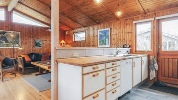 Interior acolhedor de uma cabana, com decoração em madeira, área de refeições com mesa e cadeiras, cozinha com ilha central e uma grande janela que traz luz natural.