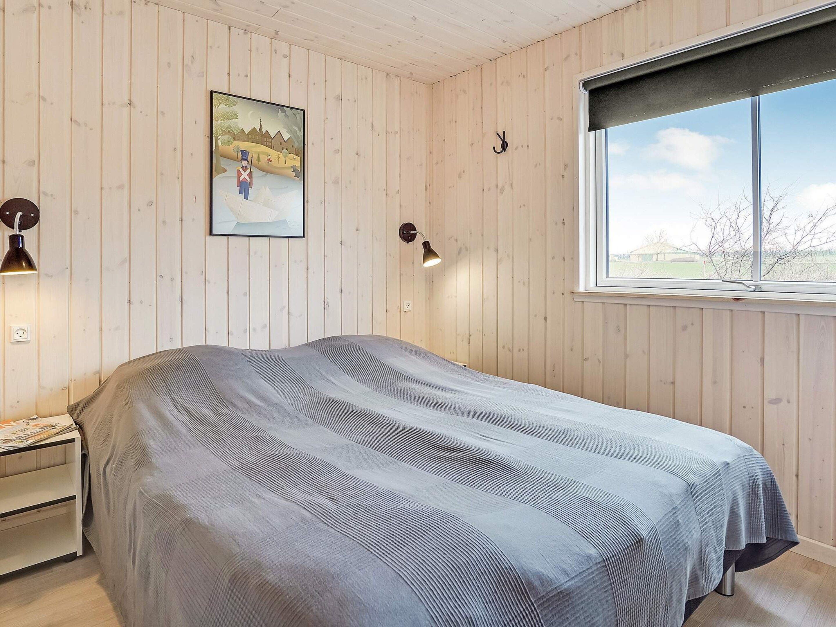 Quarto com cama, mesa de cabeceira, luminária na parede, quadro e janela.