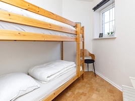 Uma cama beliche com roupa de cama branca, uma cadeira de madeira, uma pequena janela com uma planta e um chão ladrilhado.