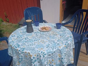 Uma mesa redonda com toalha florida, duas cadeiras azuis, um prato de biscoitos, uma caneca azul e um jarro cinza.