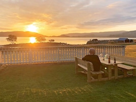 Uma pessoa sentada num banco observando o pôr do sol sobre a água, com montanhas ao longe.