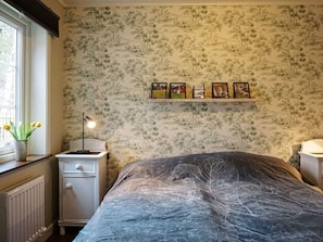 Quarto com cama, mesa de cabeceira, abajur, radiador, janela com vista e parede com quadros.