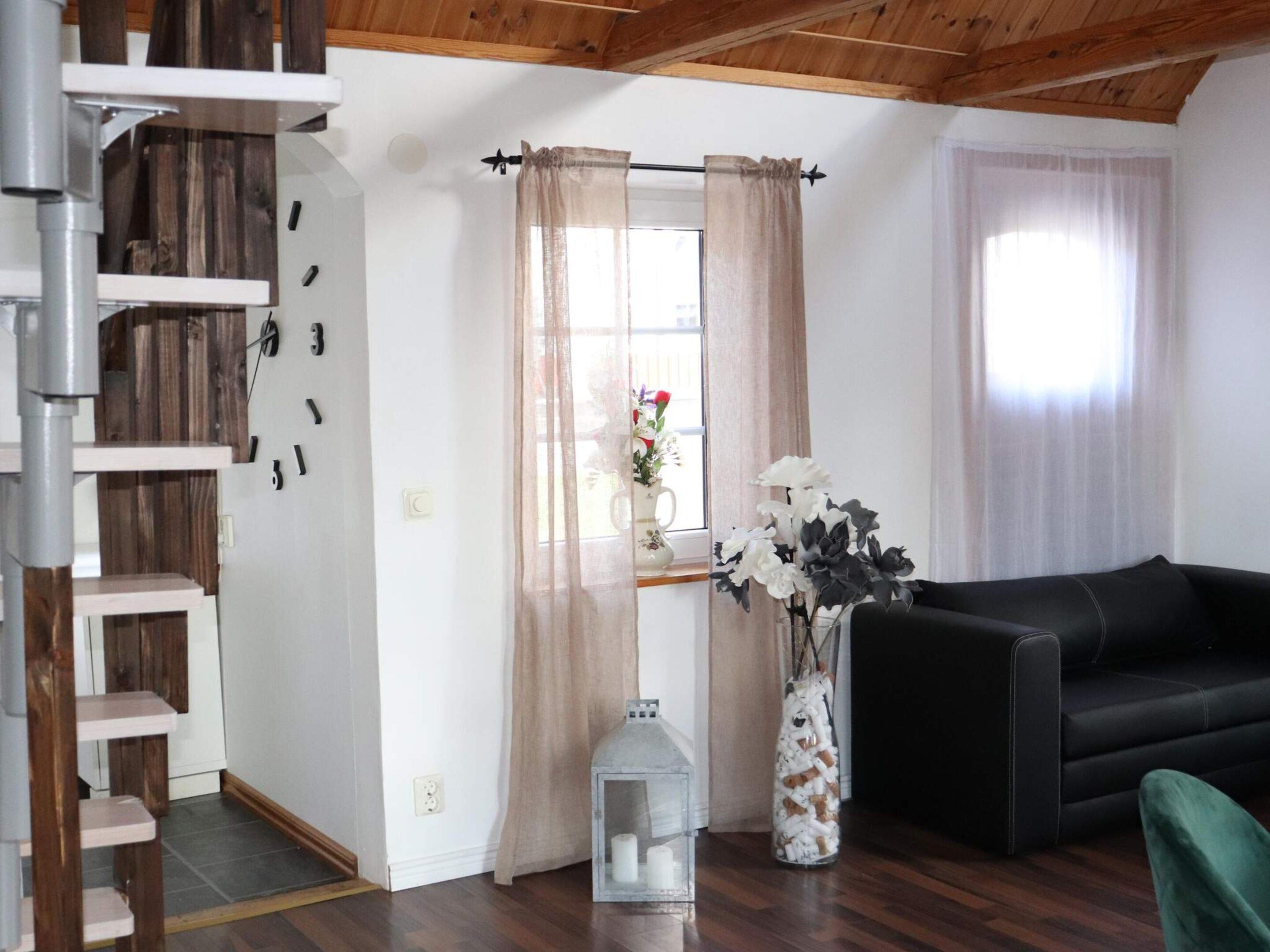 Sala de estar com sofá preto, escada de madeira e janela com cortinas finas.