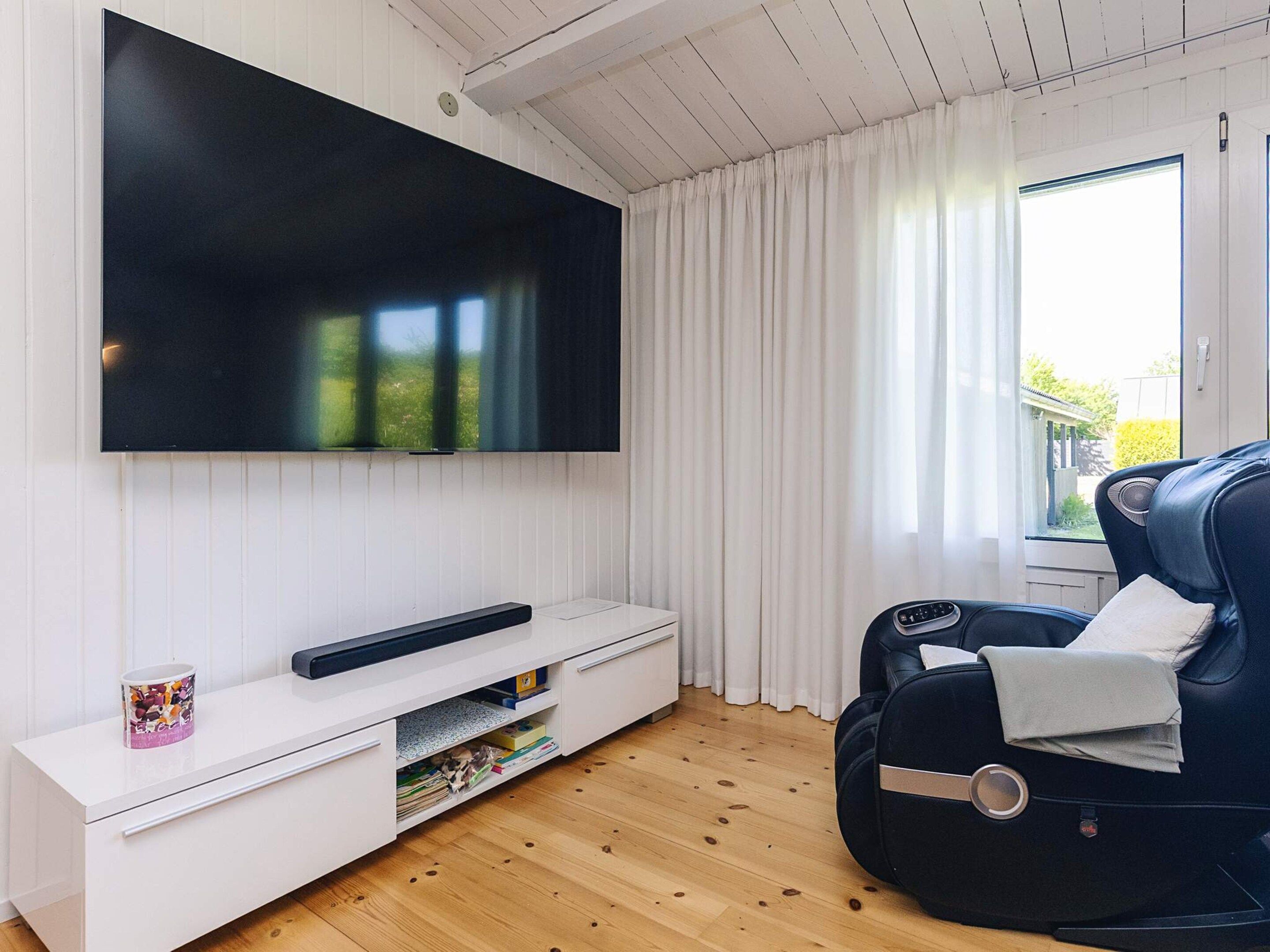 Uma sala de estar moderna com TV de tela plana fixada na parede, um rack branco, um sofá de couro e uma janela grande com cortinas.