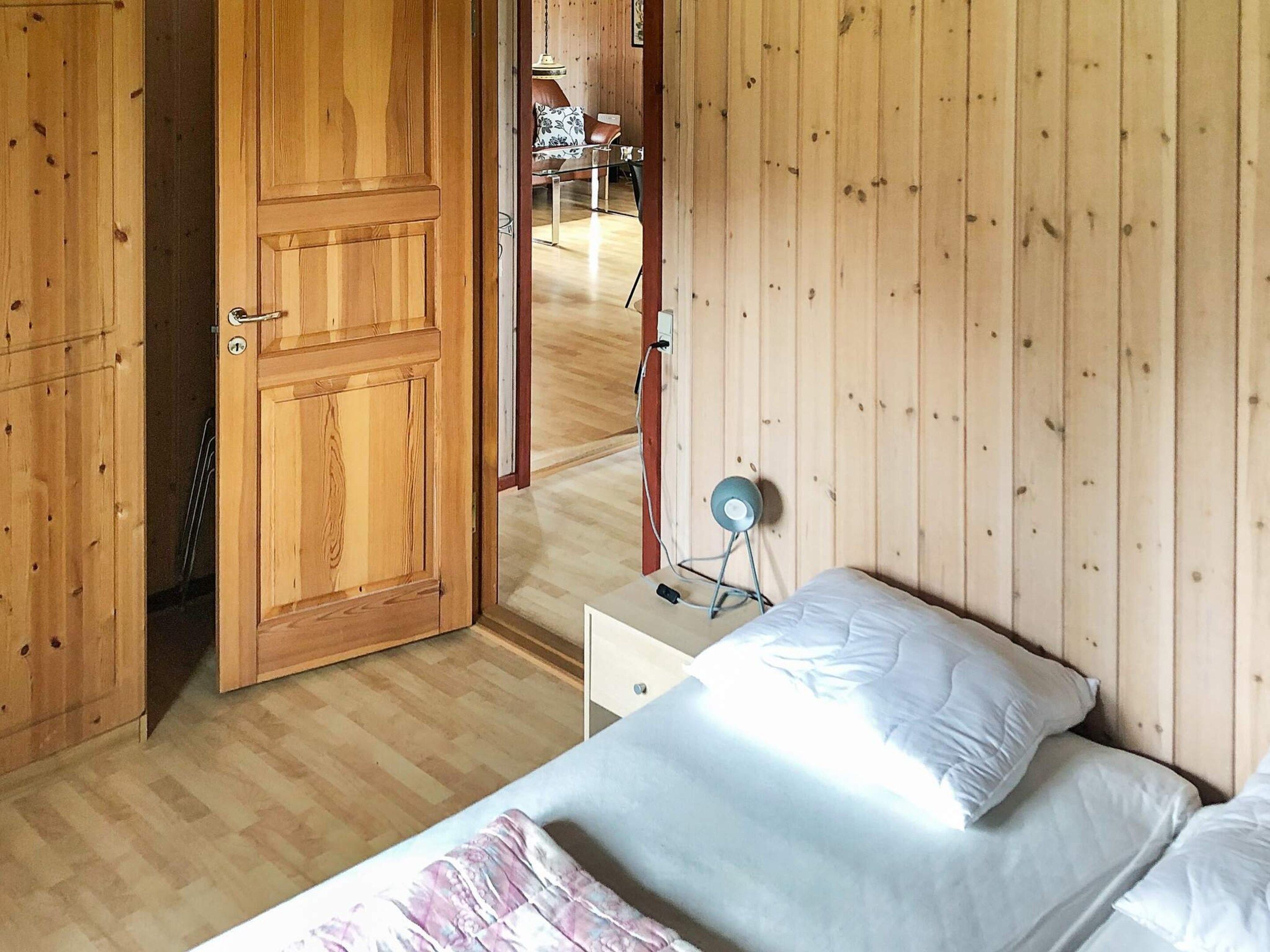 Quarto com painéis de madeira, uma cama, uma mesinha de cabeceira e uma porta que dá para outro cômodo.