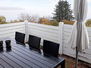 Um pátio com mesa preta e três cadeiras de vime, cerca branca e uma árvore grande ao fundo.