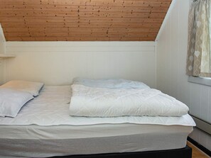 Uma cama de solteiro com roupa de cama branca em quarto com vigas de madeira no teto e paredes brancas.