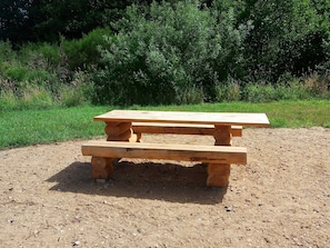 Uma mesa de piquenique de madeira com um banco, colocada em uma área de cascalho com grama e arbustos ao fundo.
