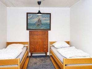Um quarto pequeno e simples, com duas camas de madeira, uma cômoda de madeira e um quadro de um veleiro na parede.