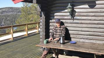 Uma pessoa sentada à mesa de madeira num terraço com vista para uma cadeia montanhosa coberta de floresta.