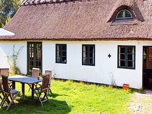 Uma casa de campo branca com telhado de palha, um pequeno jardim e uma mesa com cadeiras na parte externa.