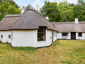 Uma casa tradicional com telhado de palha e fachada branca, uma janela com floreira e um banco na parte externa.
