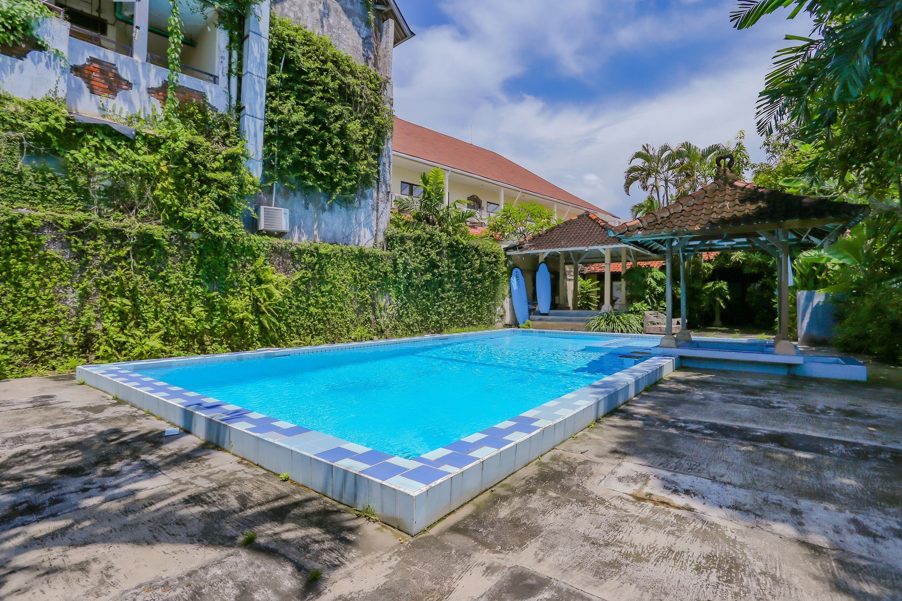 Piscina com borda em mosaico, rodeada por vegetação e um edifício com telhado vermelho.