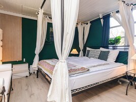 Uma cama com dossel e colunas, uma mesa de cabeceira com luminária, uma cabeceira de madeira e vista para o exterior pela janela.