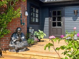 Uma estátua de uma figura sentada em um jardim com muro de tijolos, deck de madeira e uma casa com porta e janelas escuras.