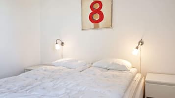 Cama bem arrumada com edredom branco e travesseiros, duas luminárias de cabeceira, uma obra de arte na parede com o número 8 e uma mesa de cabeceira branca.