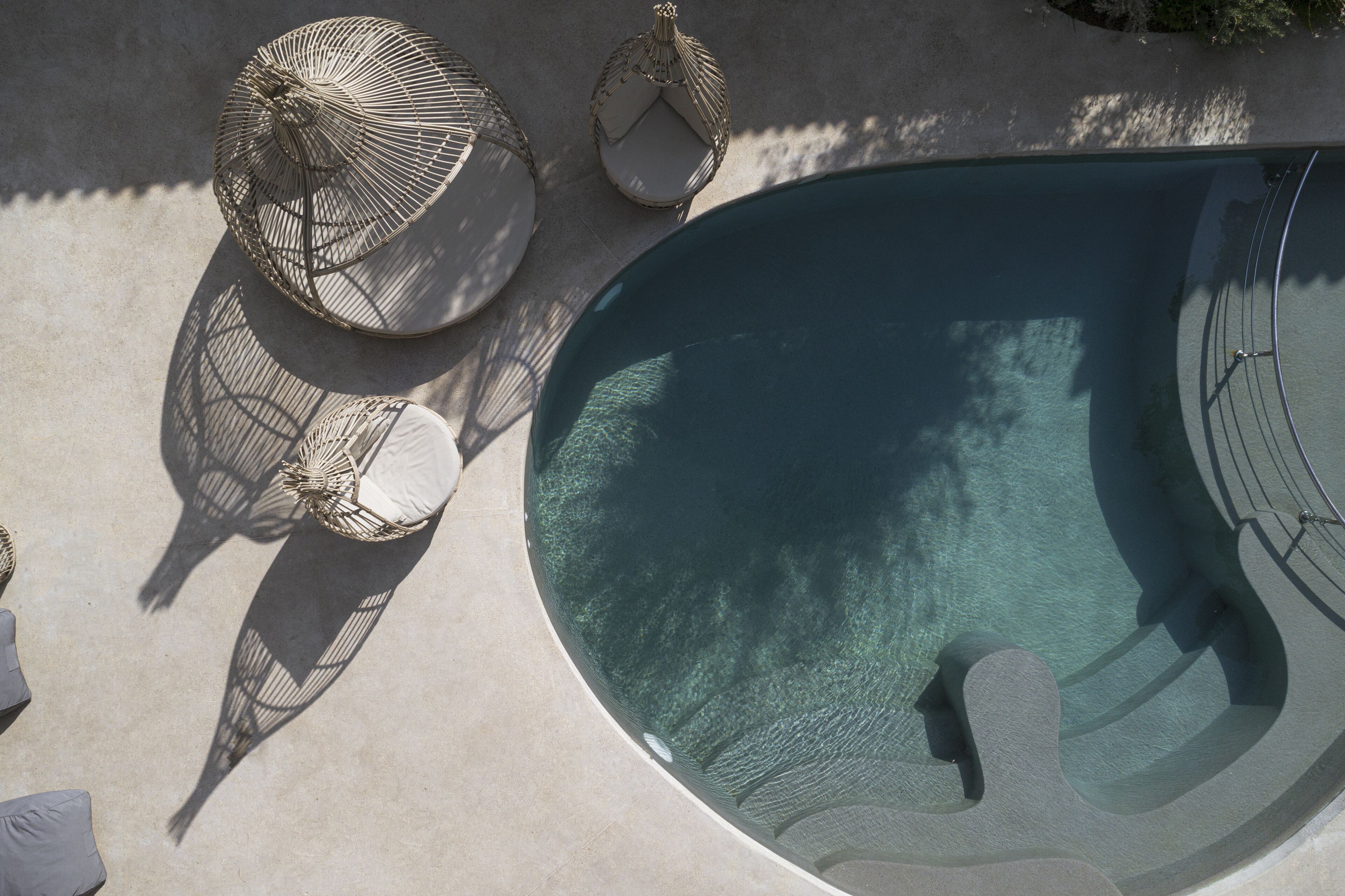 Piscina com água cristalina, rodeada por um deck de concreto. Há dois guarda-sóis de vime e alguns cadeiras de descanso.