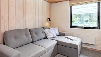 Quarto moderno com sofá seccional cinza, uma mesinha, luminária e janela com vista para área verde.