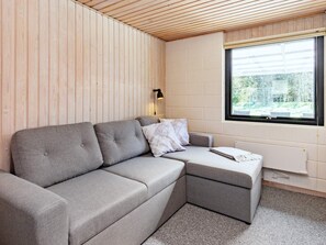 Quarto moderno com sofá seccional cinza, uma mesinha, luminária e janela com vista para área verde.