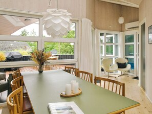 Uma sala de jantar com mesa verde, cadeiras de madeira, luminária pendente branca e um vaso com flores.