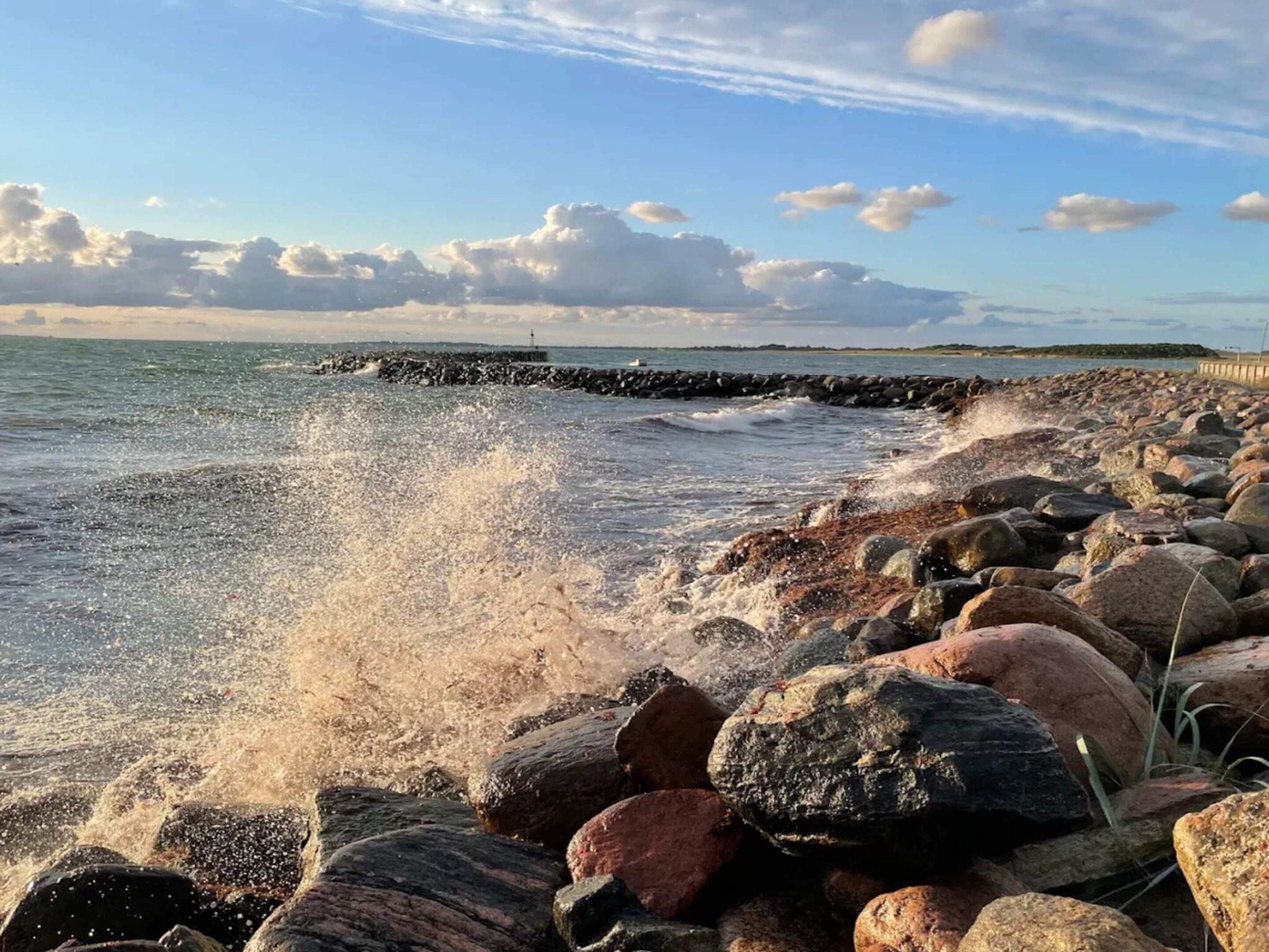 Linha costeira rochosa com ondas quebrando nas pedras e um farol ao longe.