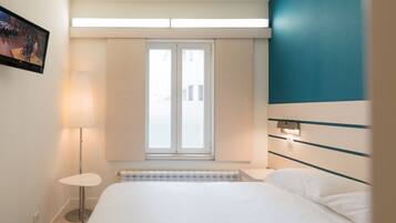 Quarto de hotel moderno com uma cama grande, uma mesinha de cabeceira, um abajur e uma janela com persianas.