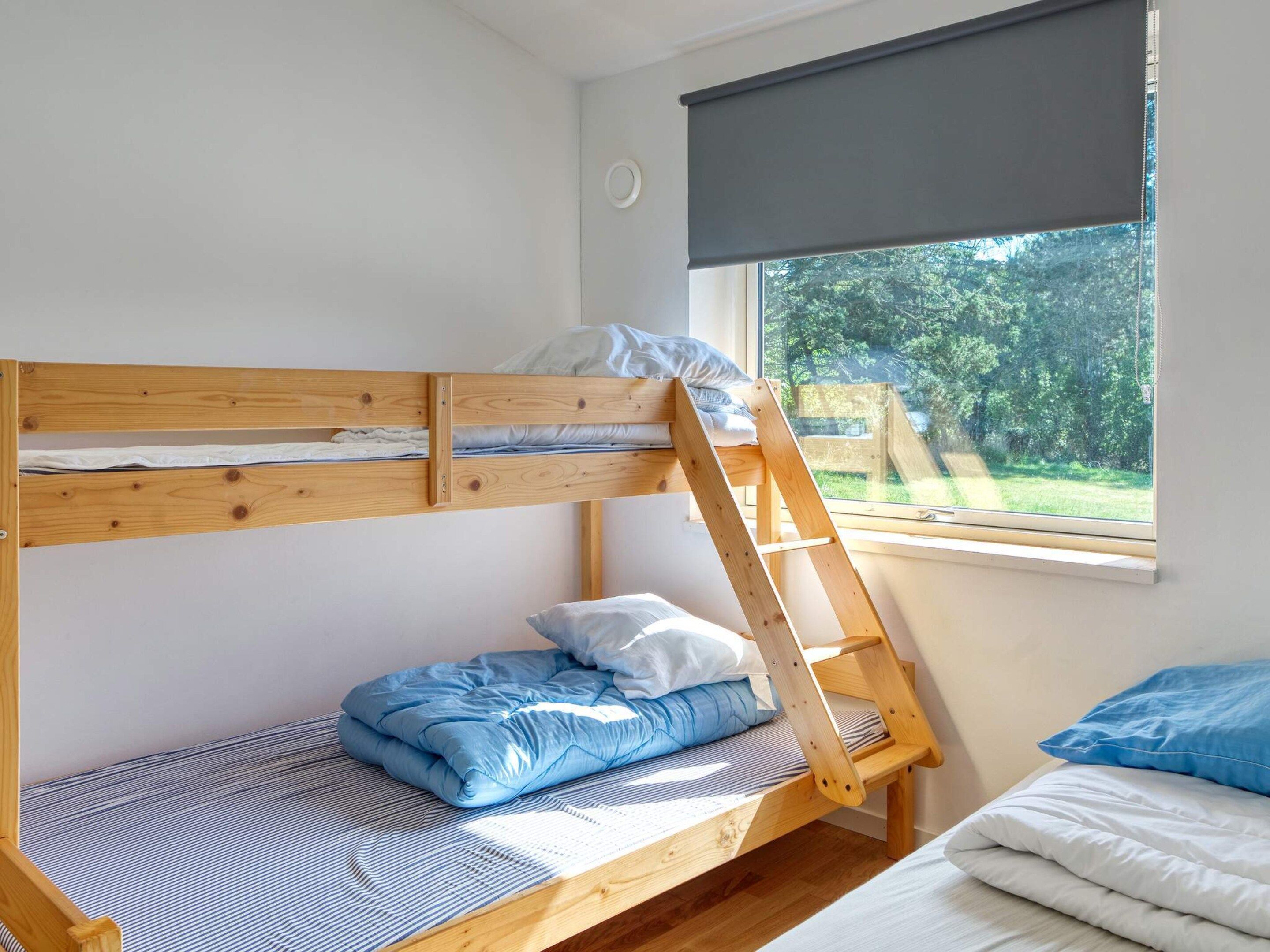 Uma cama beliche com escada de madeira, janela com vista para vegetação e uma cama com lençol listrado.