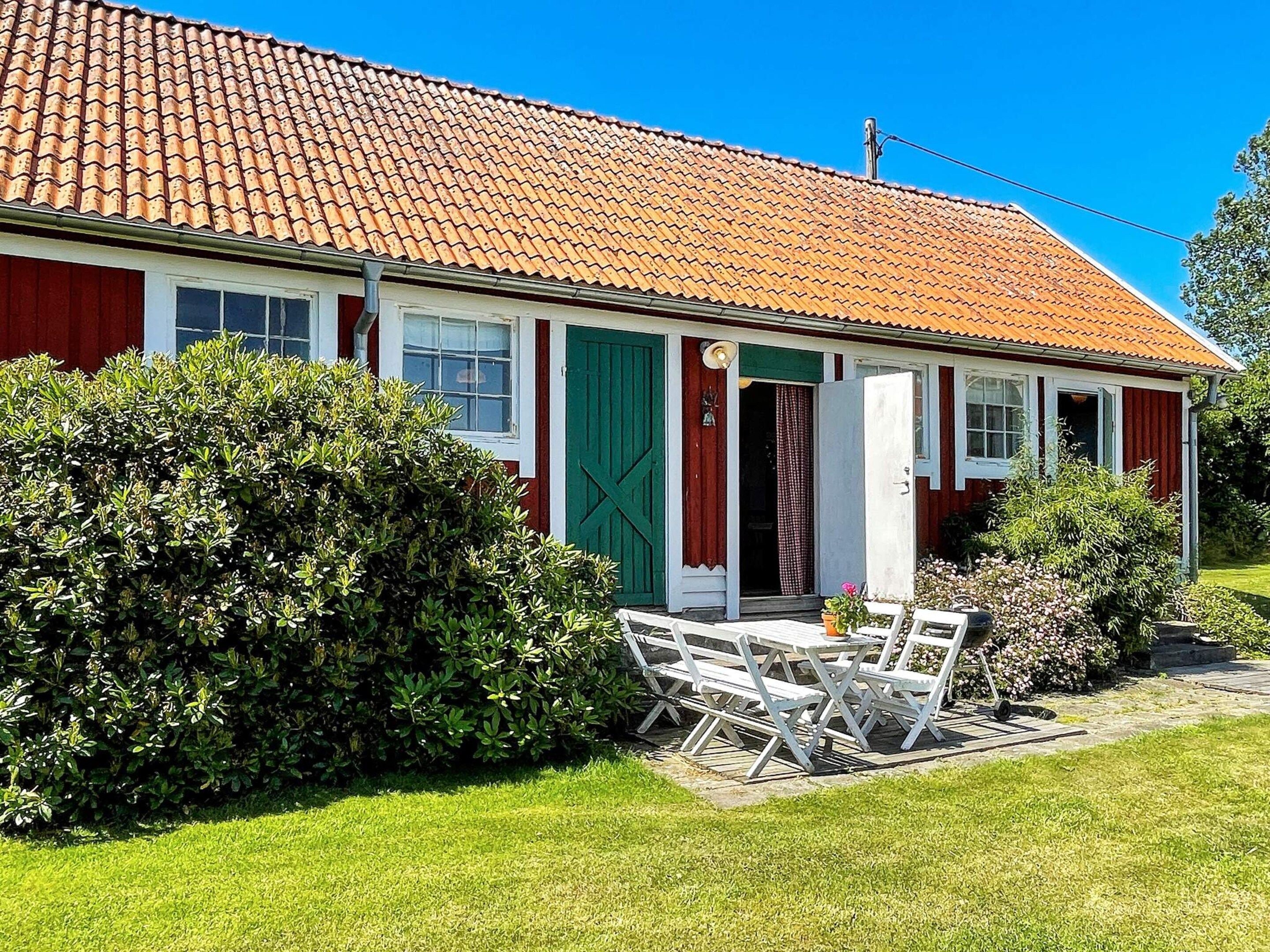 Uma casa vermelha com telhado em telhas, porta verde e um pequeno alpendre com mesa e cadeiras.