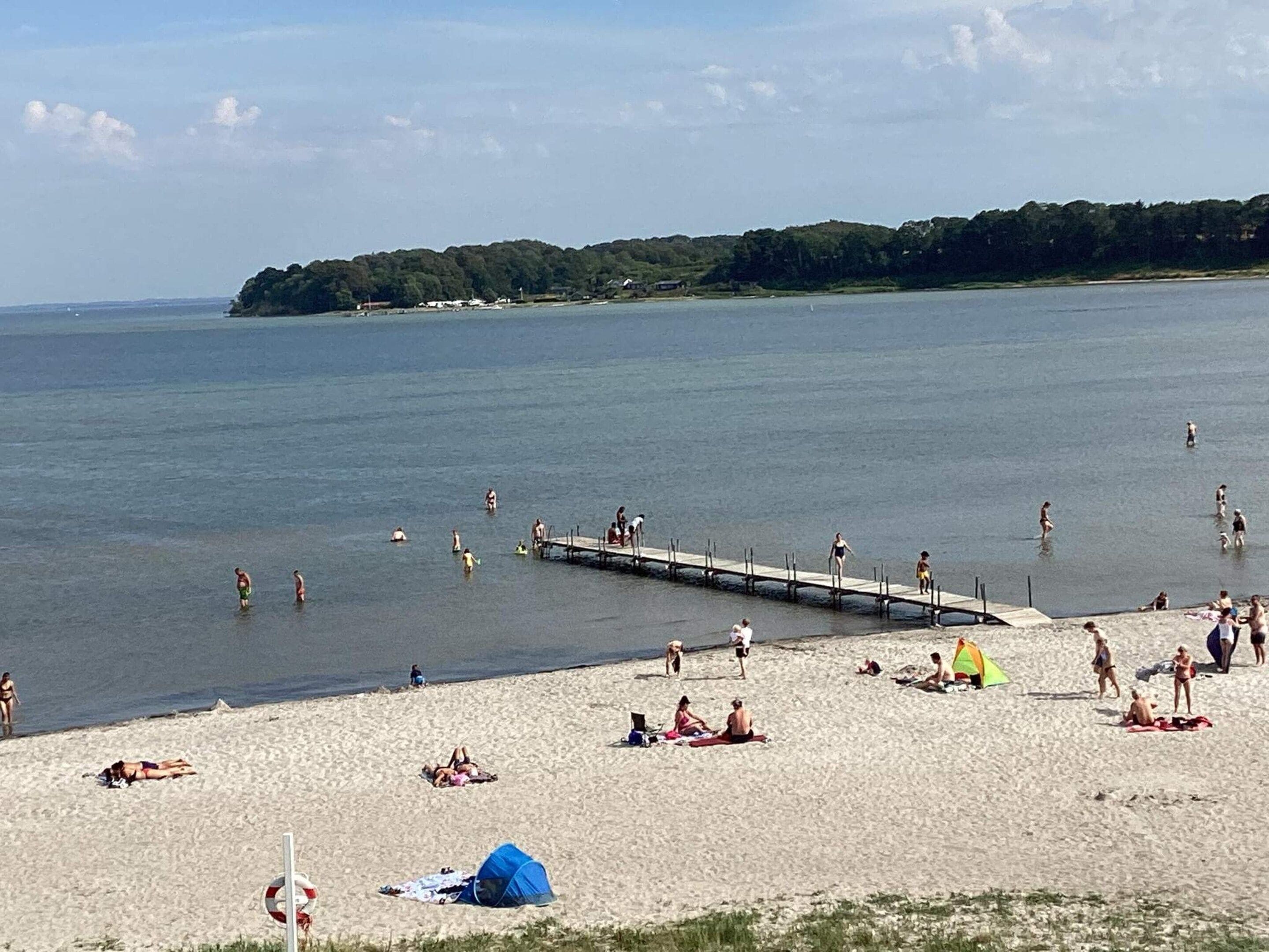 Uma praia com pessoas nadando e tomando sol, um píer que se estende na água, e uma ilha arborizada ao longe.