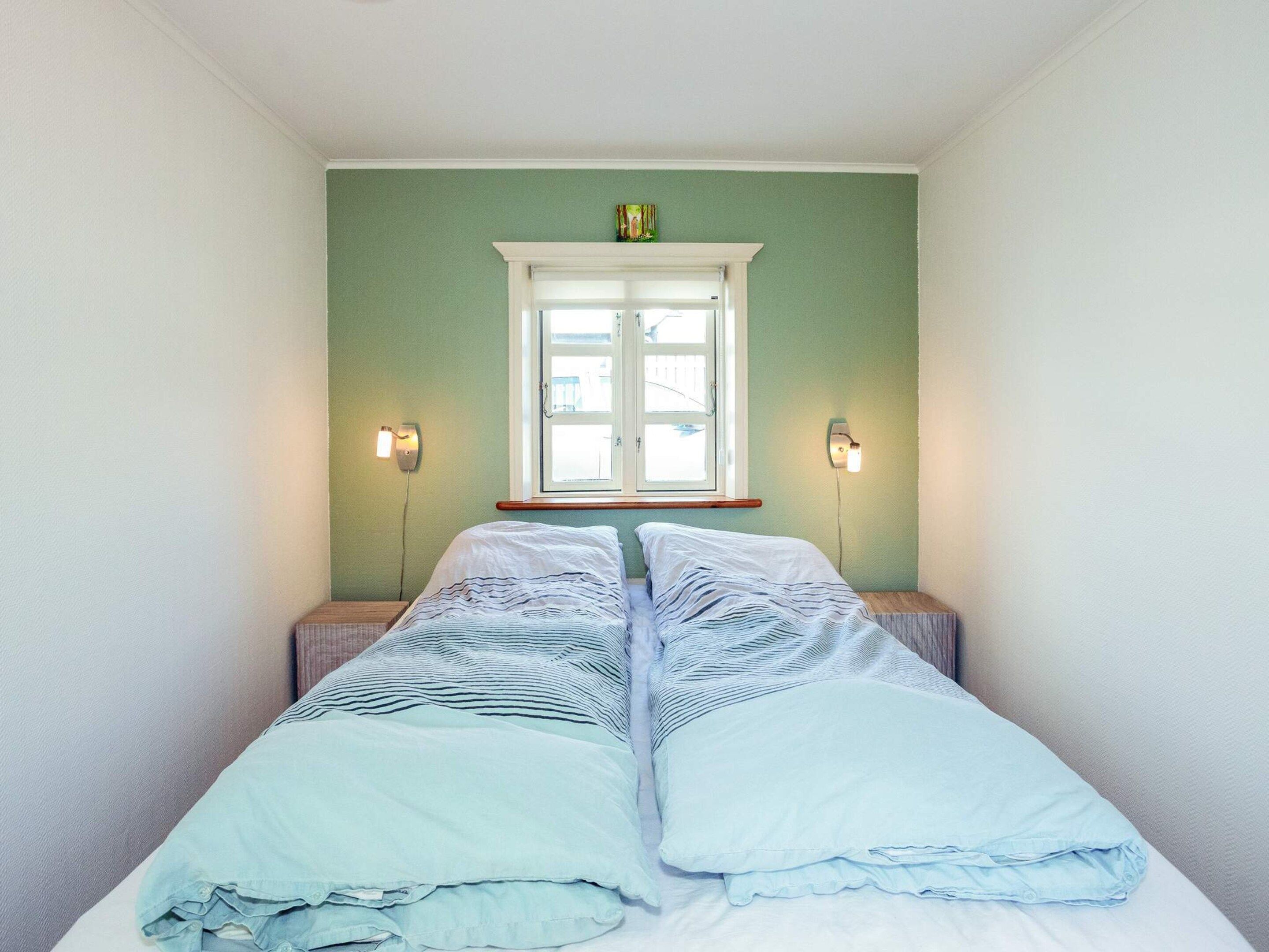 Uma cama de solteiro com colchão e dois travesseiros brancos, uma janela com moldura branca e duas luminárias fixadas na parede.