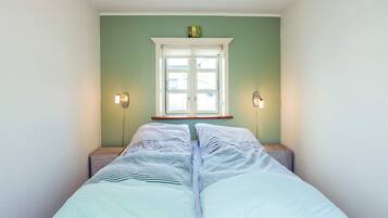 Uma cama de solteiro com colchão e dois travesseiros brancos, uma janela com moldura branca e duas luminárias fixadas na parede.