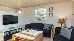 Uma sala de estar moderna com uma TV de tela plana, um sofá escuro, uma mesa de centro de madeira e uma janela com vista para árvores.