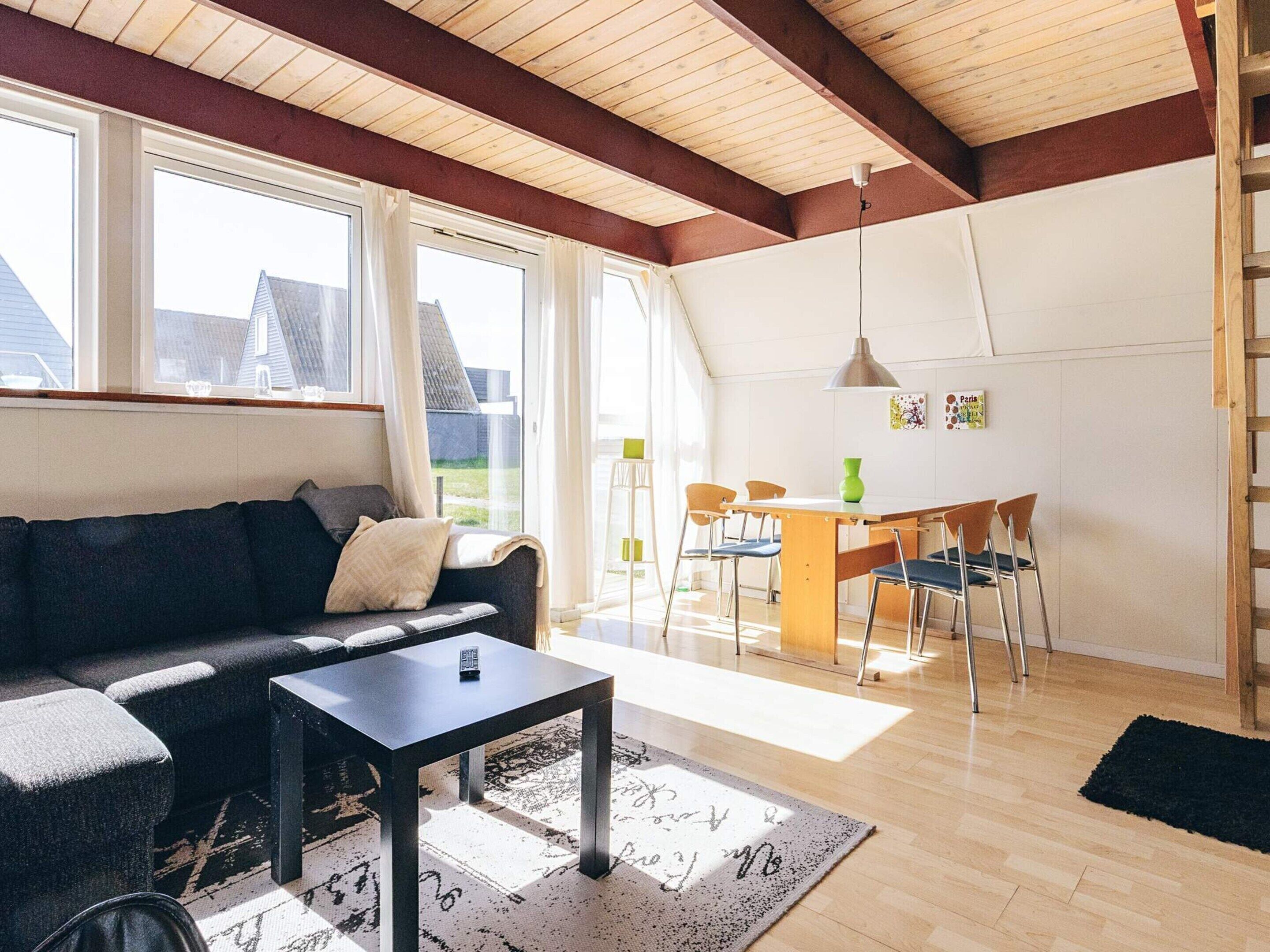 Uma sala de estar espaçosa com sofá, mesa de centro e mesa de jantar com cadeiras. Grandes janelas permitem entrada de luz natural e oferecem vista para o exterior.