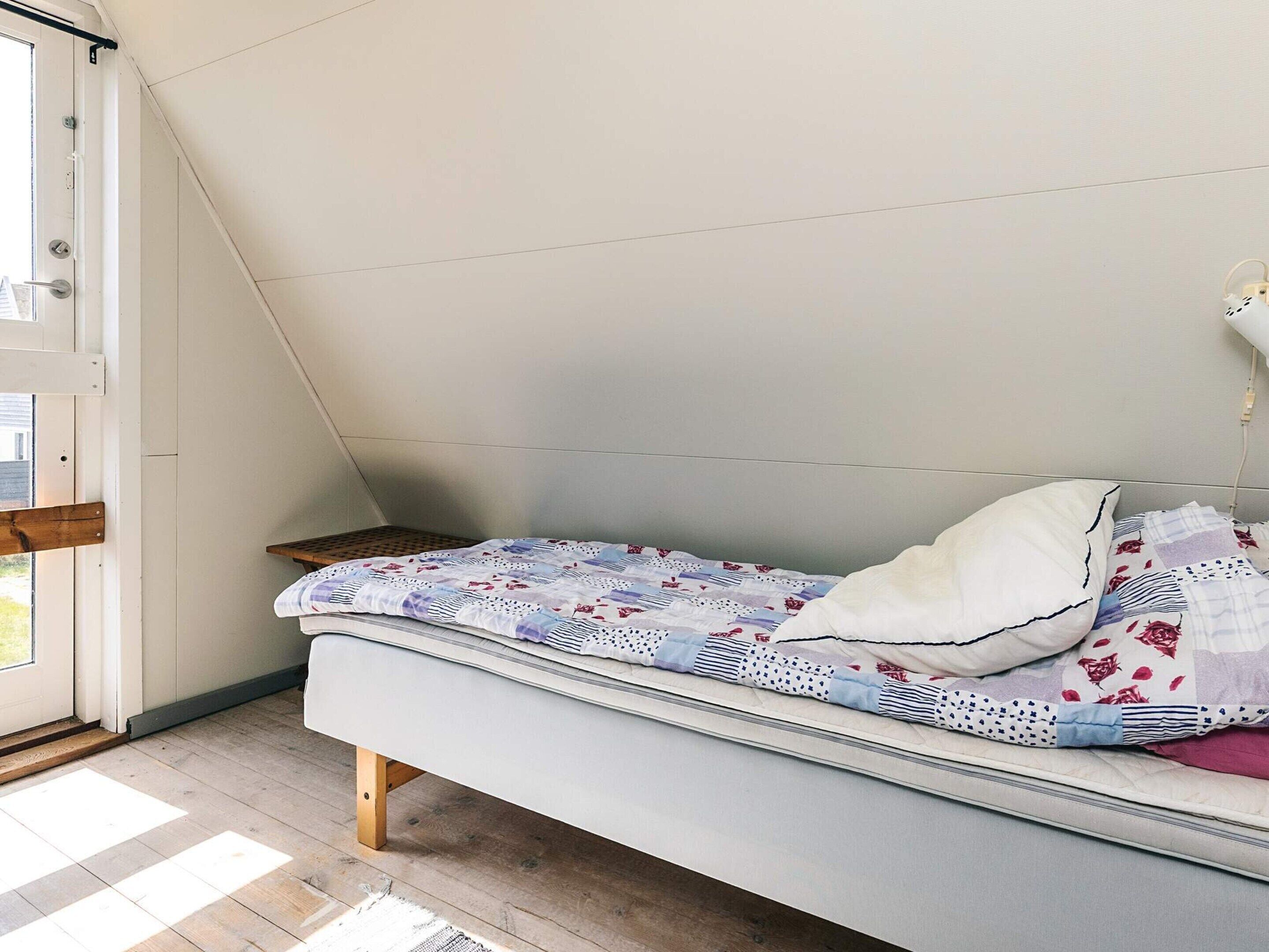 Quarto pequeno com uma cama de solteiro, teto inclinado, piso de madeira e janela com vista para o exterior.