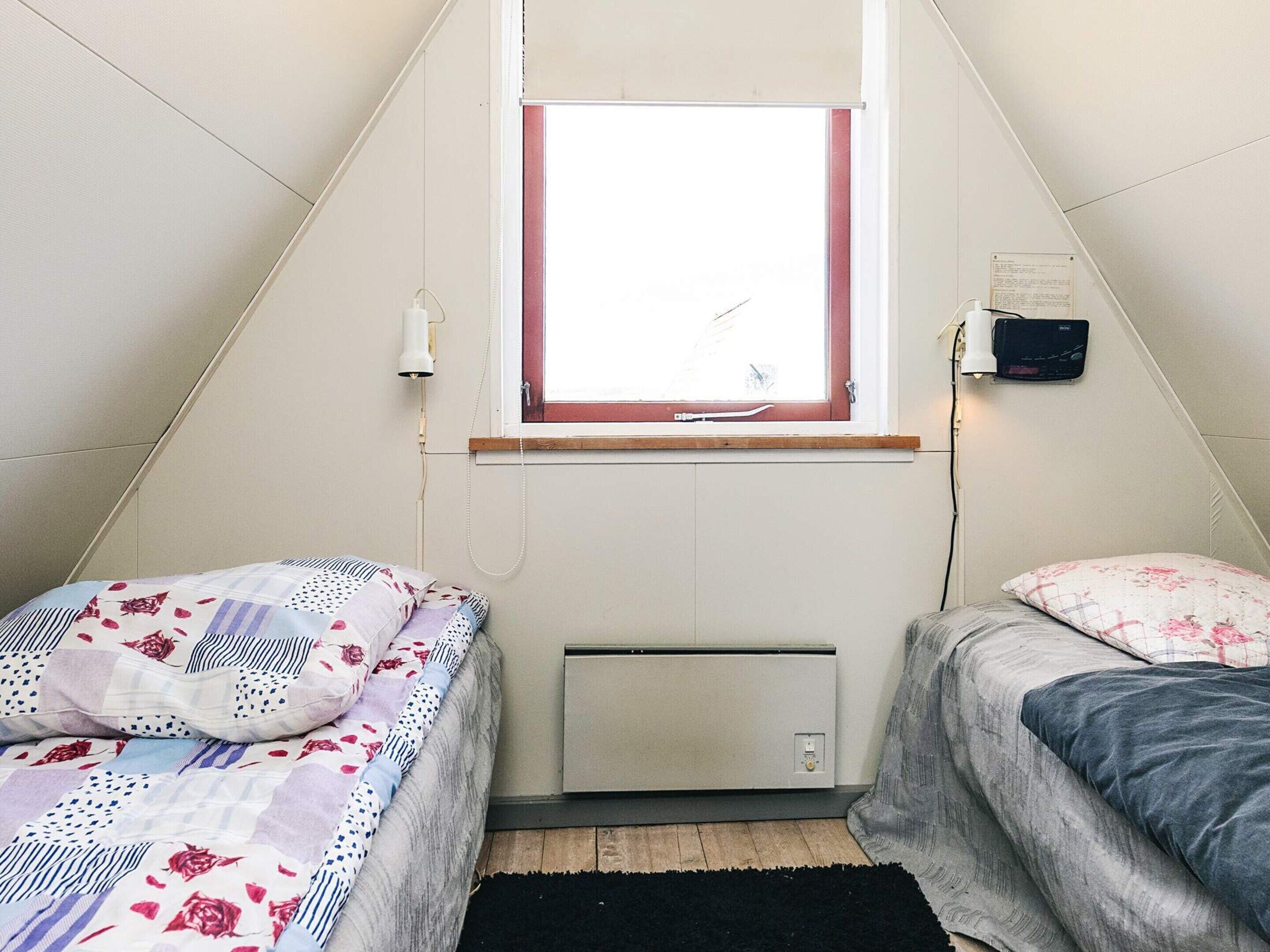 Quarto pequeno em formato triangular com duas camas, uma janela com moldura vermelha, aquecedor fixado na parede e um pequeno prateleira.