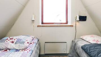 Quarto pequeno em formato triangular com duas camas, uma janela com moldura vermelha, aquecedor fixado na parede e um pequeno prateleira.