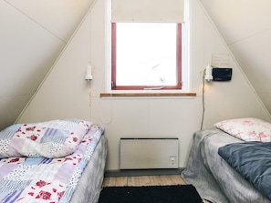 Quarto pequeno em formato triangular com duas camas, uma janela com moldura vermelha, aquecedor fixado na parede e um pequeno prateleira.