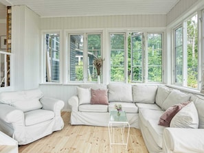 Quarto com sofá branco, uma mesa pequena e janelas amplas com vista para o verde.