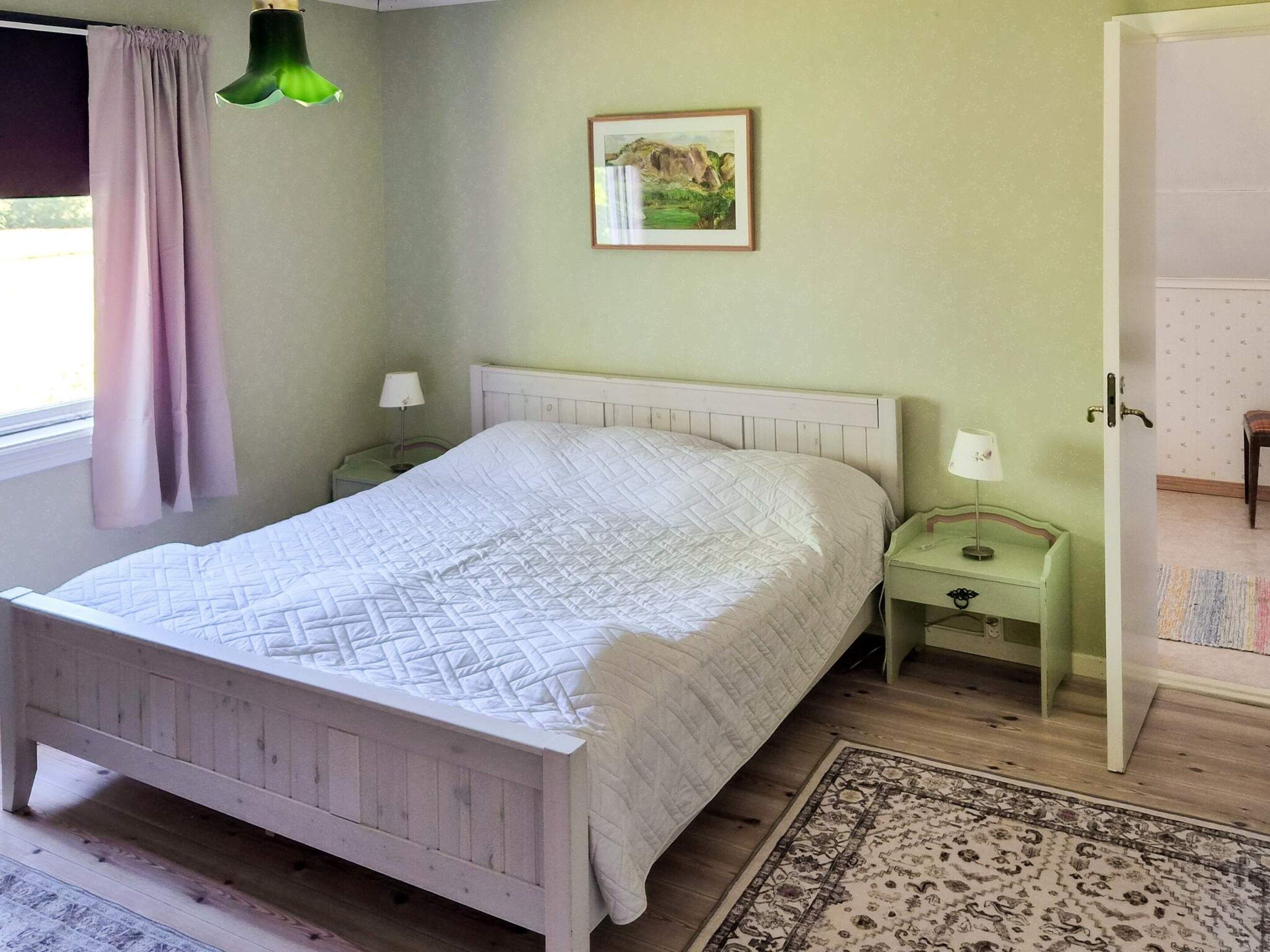 Quarto com cama, mesas de cabeceira, luminária, quadro na parede e porta para outro cômodo.