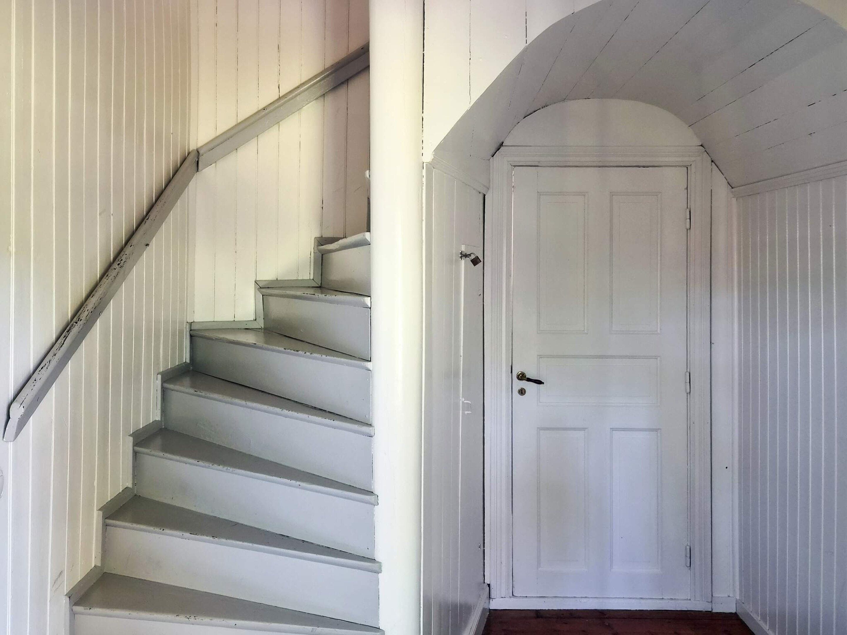 Escada de madeira branca que leva a uma porta branca com corrimão de metal.