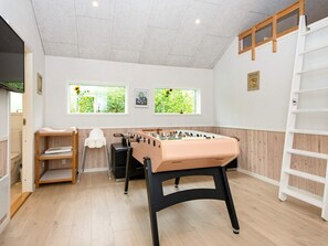 Quarto com mesa de futebol, prateleira branca, escada e janelas com vegetação lá fora.