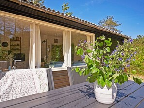 Uma área de estar ao ar livre aconchegante, com uma mesa, um vaso de planta e vista para uma casa com janelas e cortinas grandes.