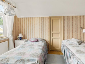 Quarto com duas camas, mesa de cabeceira, luminária, janela com cortinas e porta.