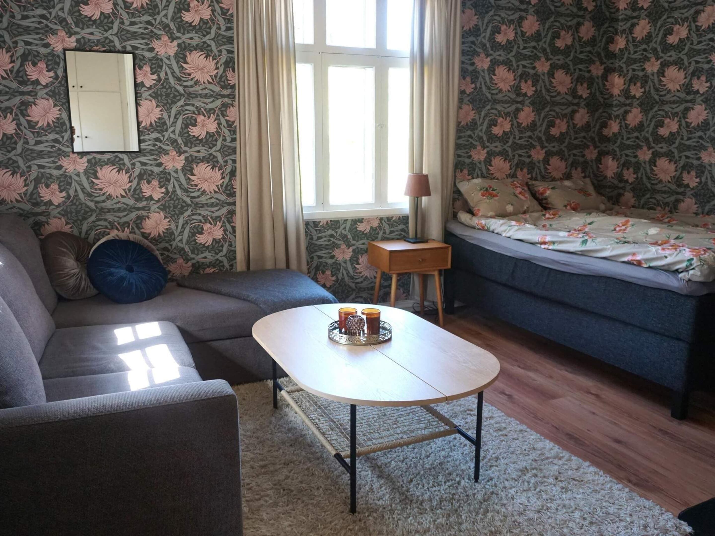 Quarto com sofá, mesinha com bandeja, luminária e cama com roupa de cama florida.