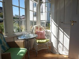 Um quarto iluminado pelo sol, com uma cadeira de vime, uma mesa pequena e uma porta branca.