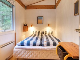 Quarto com cama, mesa de madeira, relógio fixado na parede e janela com vista para árvores.