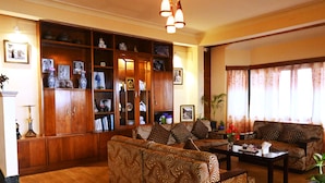 Uma sala de estar com sofás estampados, um armário de madeira com diversos objetos e uma janela grande com cortinas.