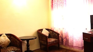 Um quarto com duas poltronas de madeira, uma mesa redonda pequena, uma televisão sobre um móvel de madeira e uma janela com cortinas florais.